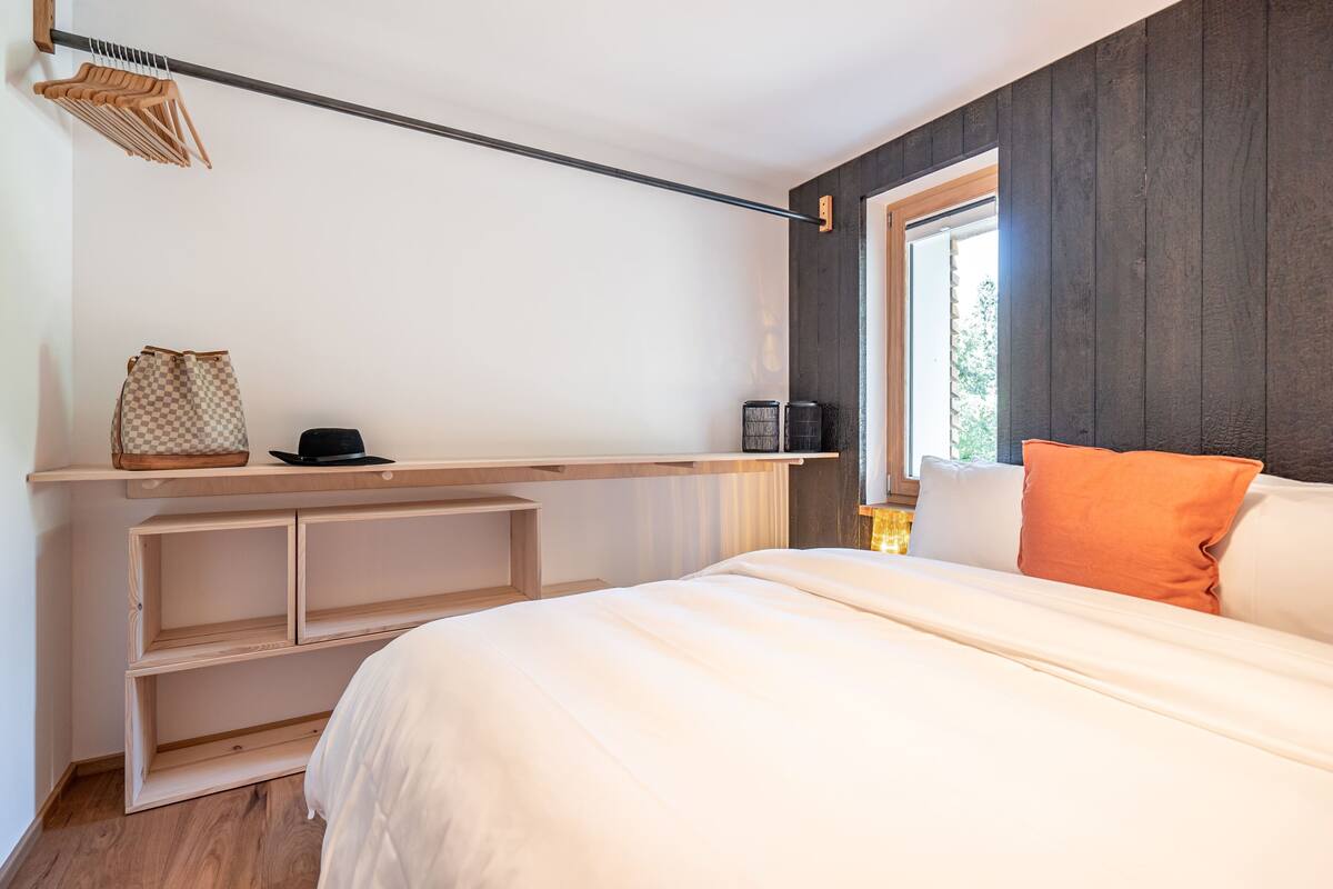 Um quarto com uma cama, uma prateleira com uma mala e um chapéu, uma janela e uma parede de madeira.