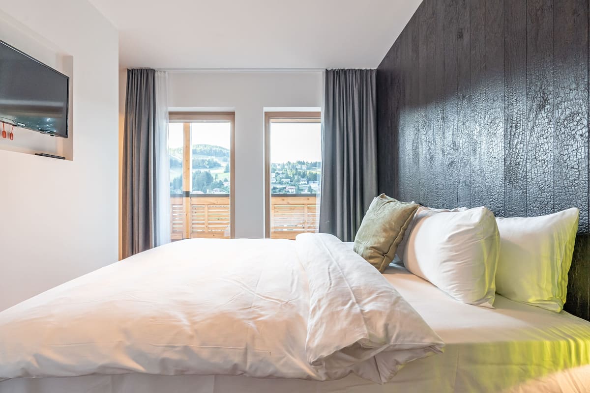 Quarto moderno com uma cama grande, duas janelas com cortinas, uma TV de tela plana na parede e vista para a cidade.