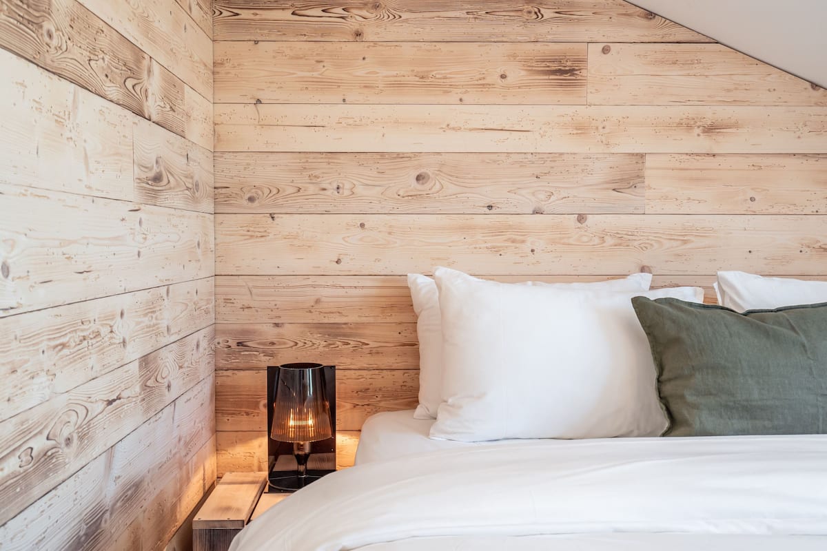 Quarto com paredes de painel de madeira, uma cama com travesseiros brancos e verdes, e uma mesa de cabeceira com uma luminária.