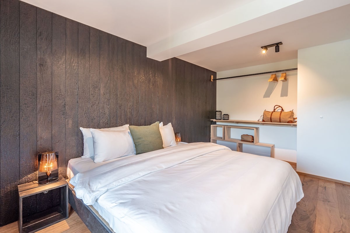Quarto moderno com uma cama grande, mesa de cabeceira com abajur e espelho sobre a cama.