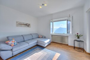 Uma sala de estar moderna com um sofá seccional cinza, um tapete azul e branco, uma pequena mesa lateral com uma planta e uma janela com vista para as montanhas.