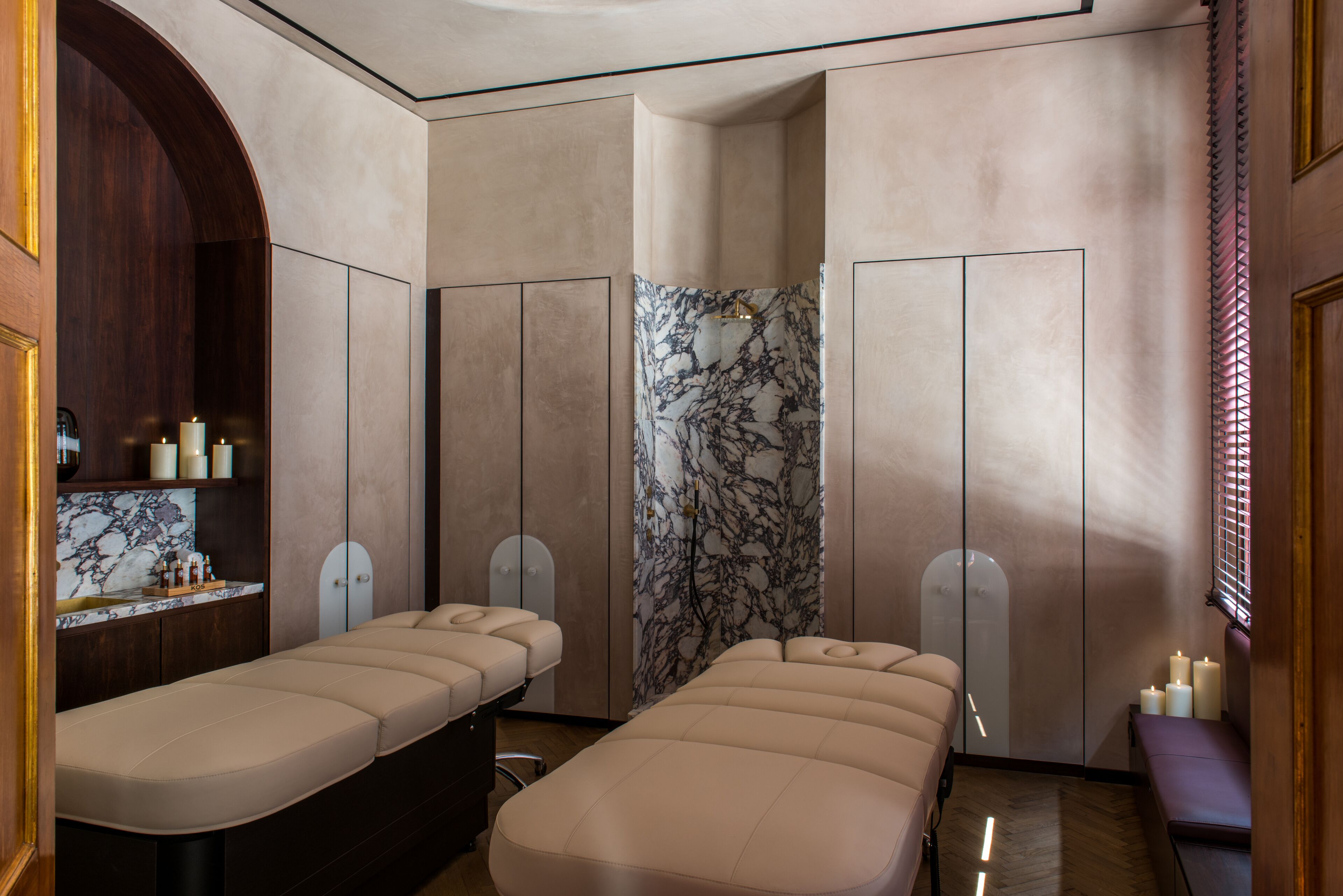 Sala de spa com duas mesas de massagem, parede de mármore e toucador de madeira com velas.