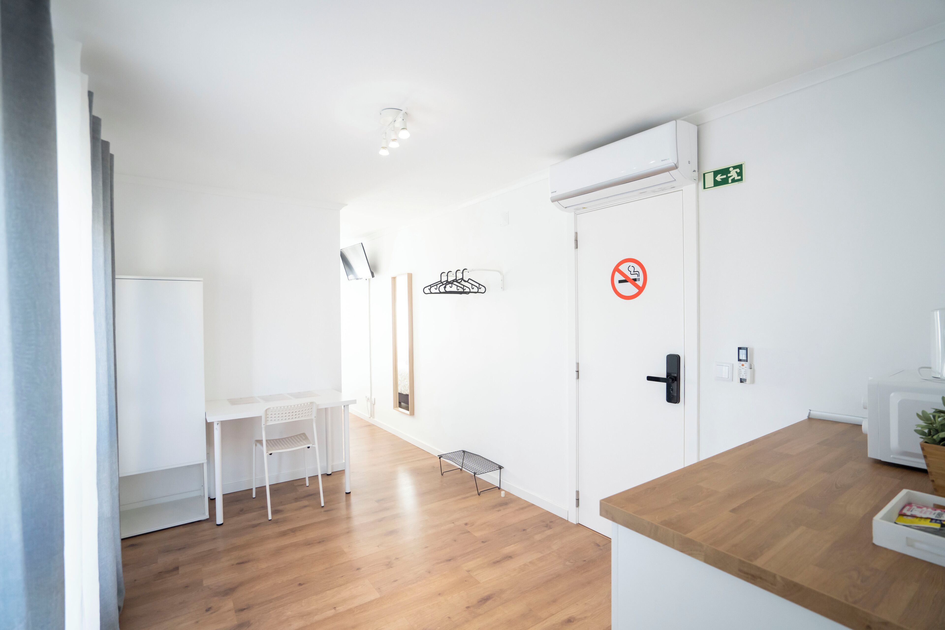 Quarto limpo e moderno, com piso de madeira, uma escrivaninha branca, uma cadeira e um quadro branco. Há uma porta com um aviso de proibido fumar e sinal de saída.