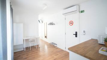 Quarto limpo e moderno, com piso de madeira, uma escrivaninha branca, uma cadeira e um quadro branco. Há uma porta com um aviso de proibido fumar e sinal de saída.