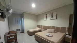 Quarto de hotel com uma cama grande, uma escrivaninha e uma cadeira. Há um ventilador de teto e um quadro na parede.
