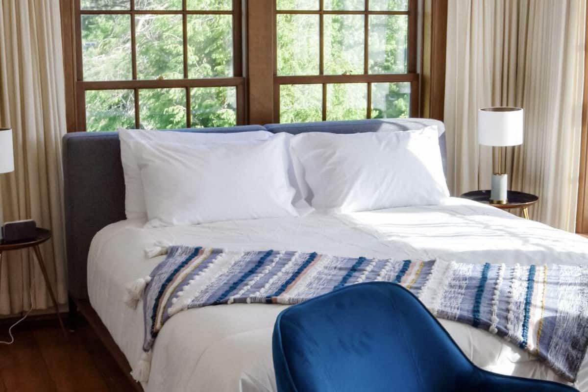 Quarto com cama, uma cadeira azul, mesa de cabeceira com abajur e janela com cortinas.