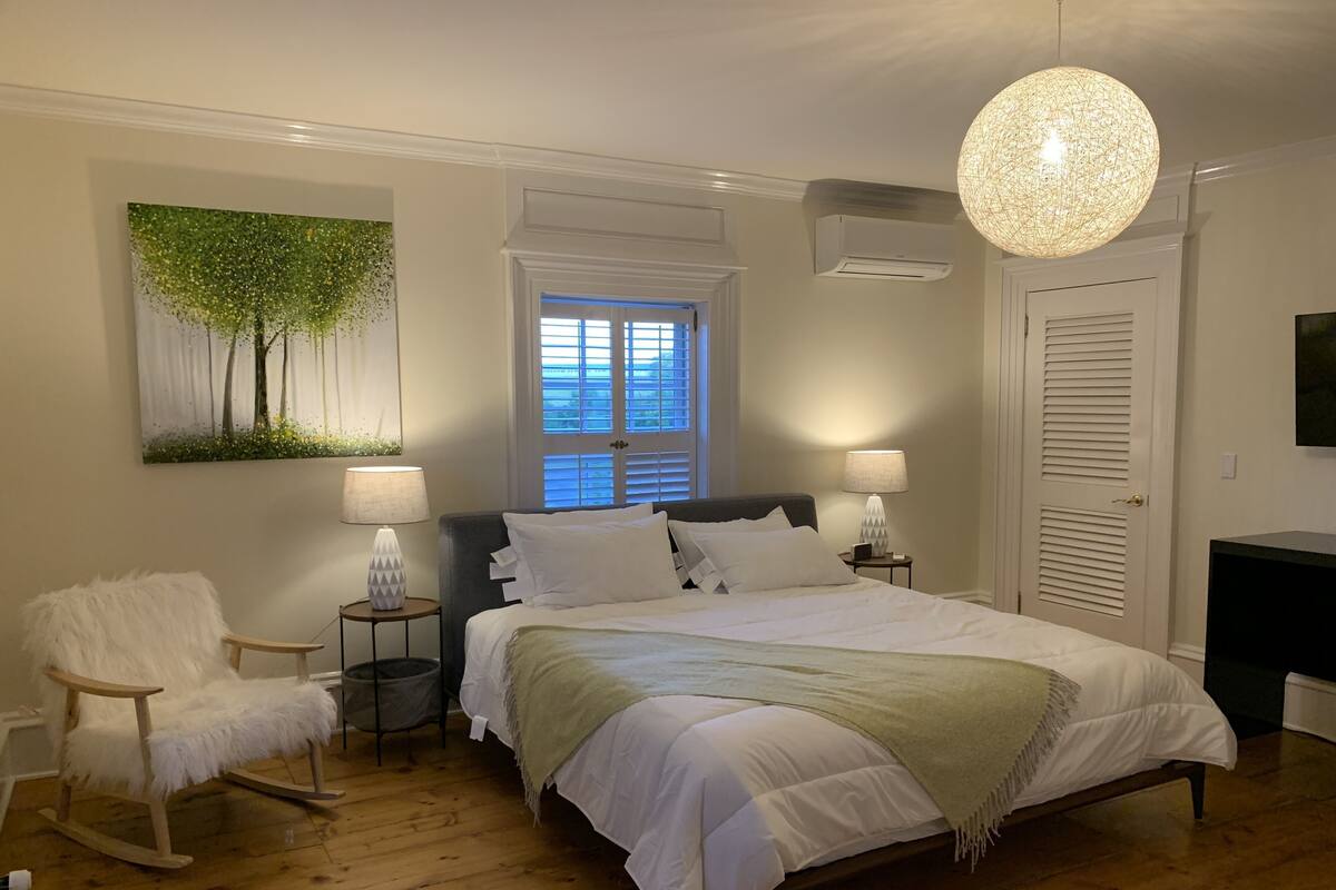 Quarto com cama, duas mesas de cabeceira, uma cadeira de balanço, janela e luminária pendente.