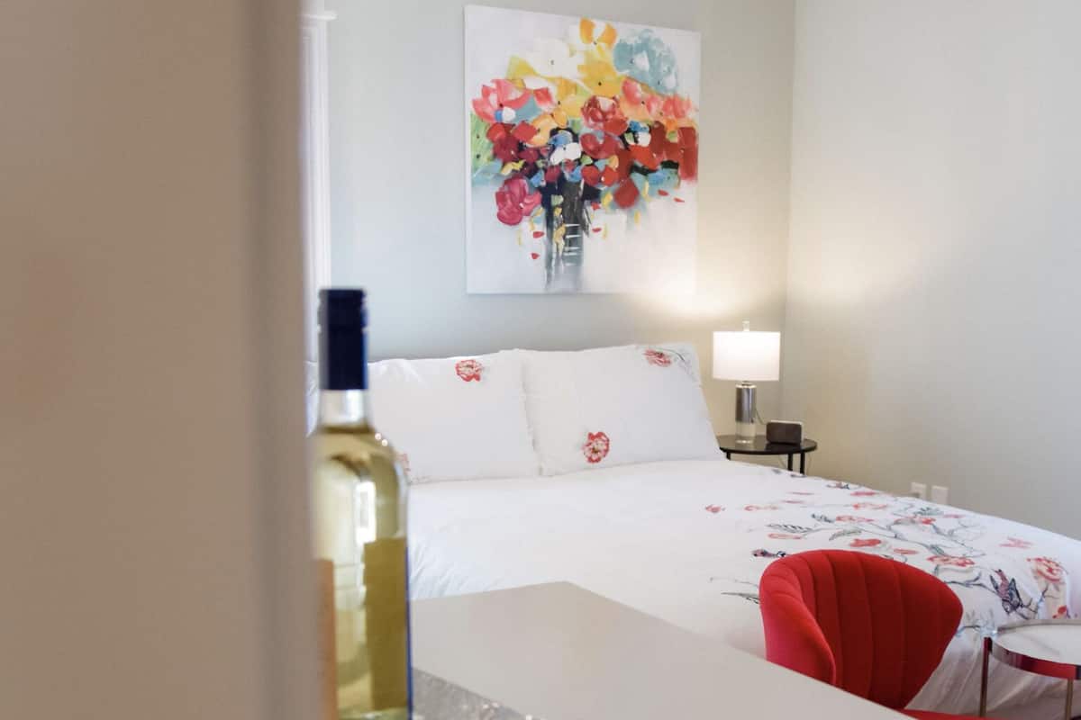 Quarto de hotel com uma cama, mesa de cabeceira, abajur, um quadro na parede e uma garrafa sobre a mesa.