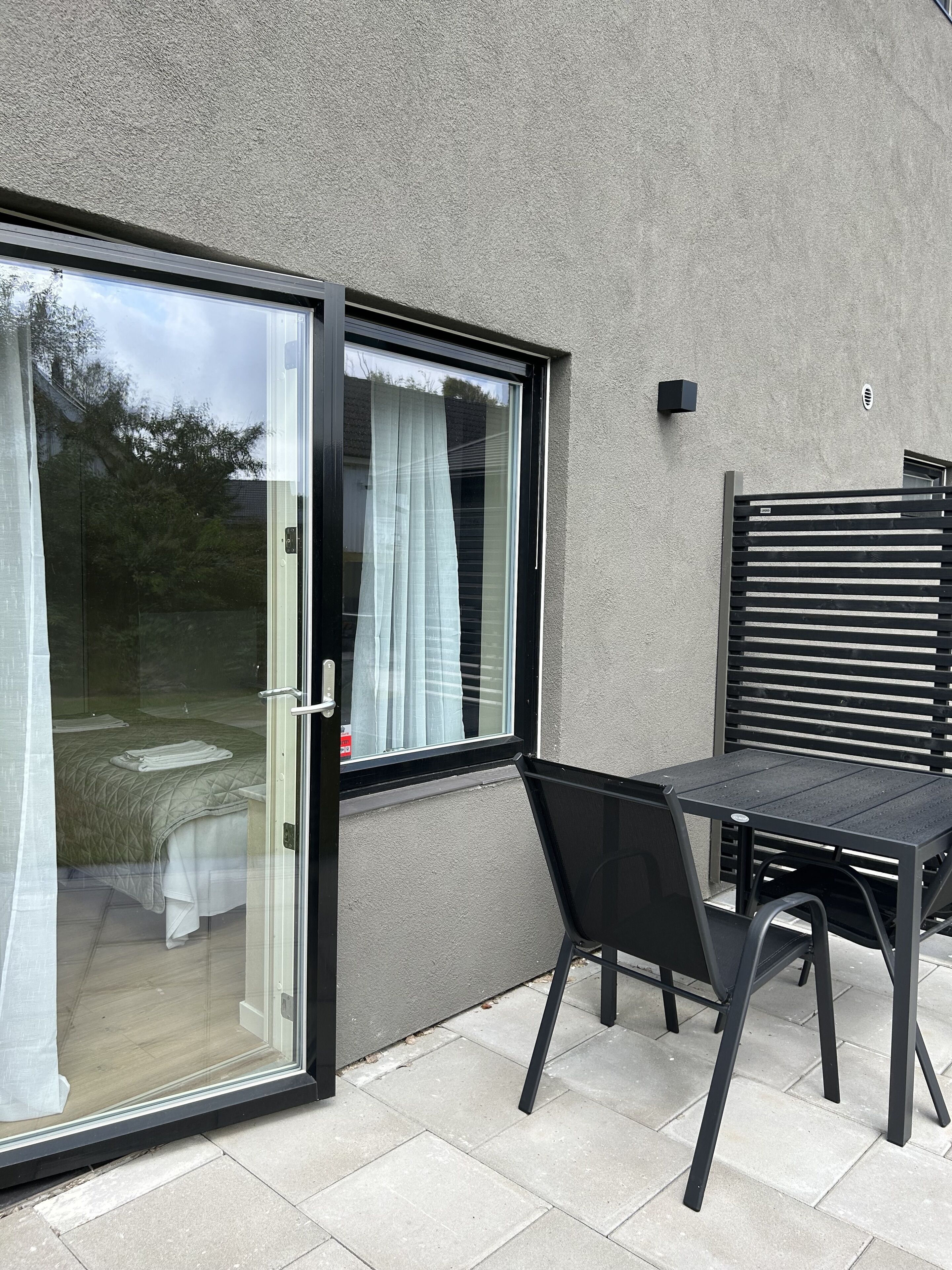Uma área de varanda moderna ao ar livre com porta de vidro dando para um quarto, mesa e cadeiras de metal preto e vista do jardim pela janela.