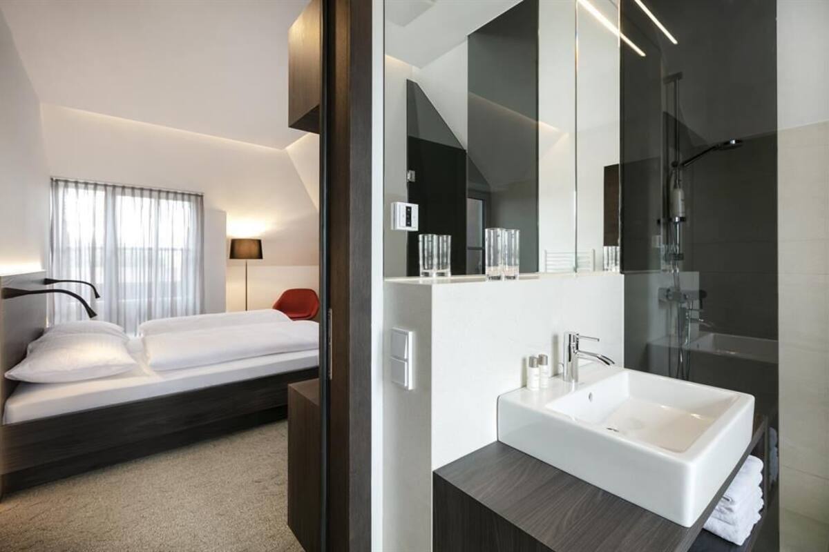 Quarto de hotel moderno com uma cama grande, banheiro separado com um espelho amplo e mesa de cabeceira com abajur.
