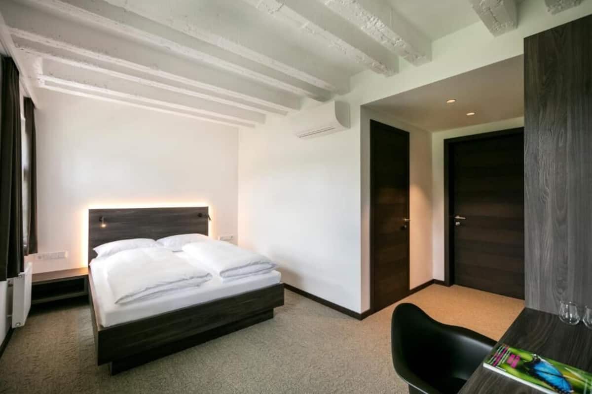 Quarto de hotel com uma cama, escrivaninha, cadeira, porta e ar condicionado fixado na parede.