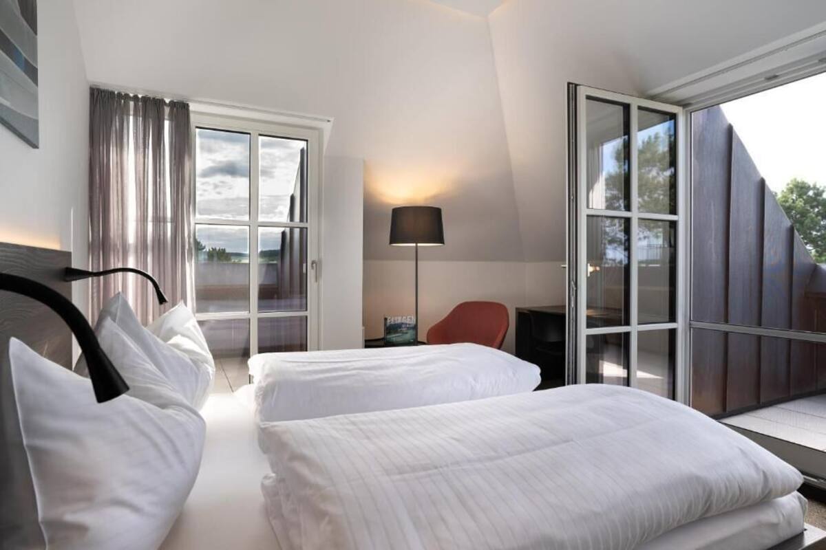 Quarto de hotel com duas camas, uma escrivaninha, uma cadeira, uma luminária e janelas amplas.