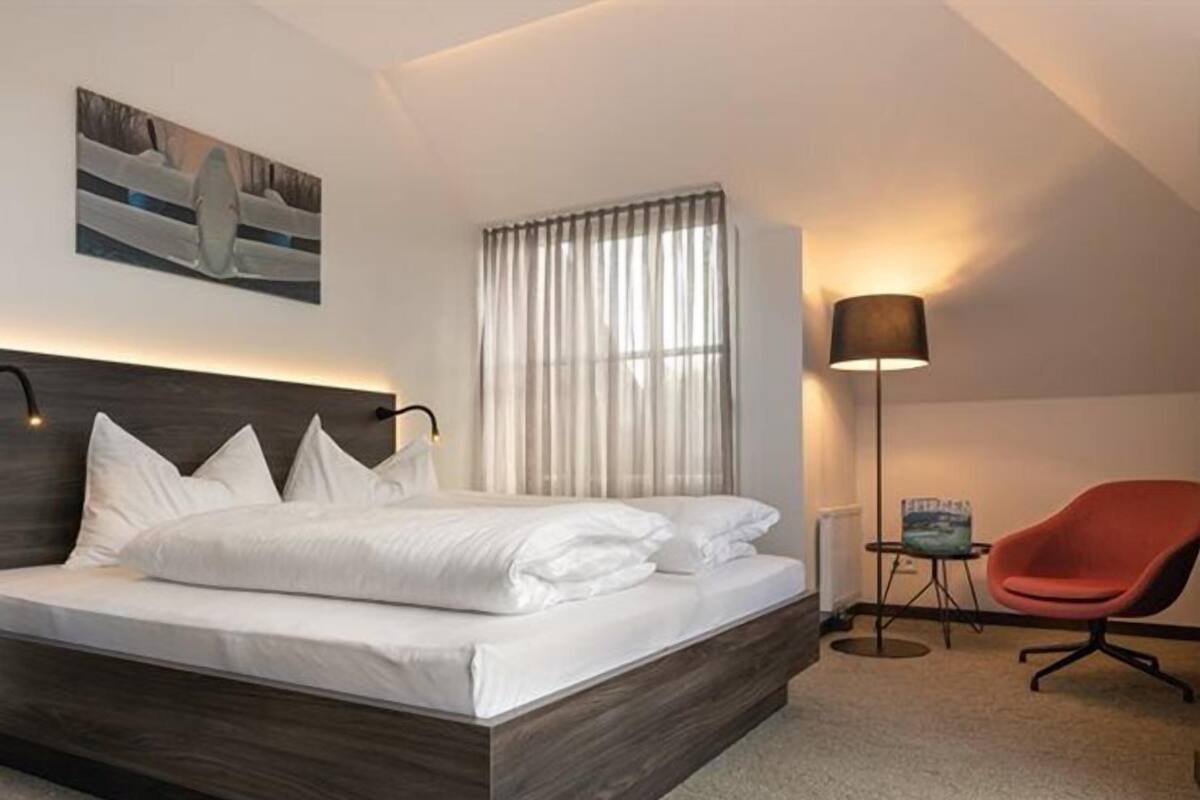 Quarto de hotel moderno com uma cama grande, uma luminária de cabeceira, uma cadeira vermelha e uma janela com cortinas.