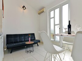 Um espaço de estar moderno com um sofá de couro preto, uma cadeira branca, uma mesa redonda pequena com uma garrafa e copos, e uma janela grande com vista.