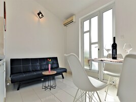Uma sala de estar moderna com um sofá de couro preto, uma cadeira branca, uma mesinha redonda e uma mesa de jantar com dois copos de vinho e uma garrafa.
