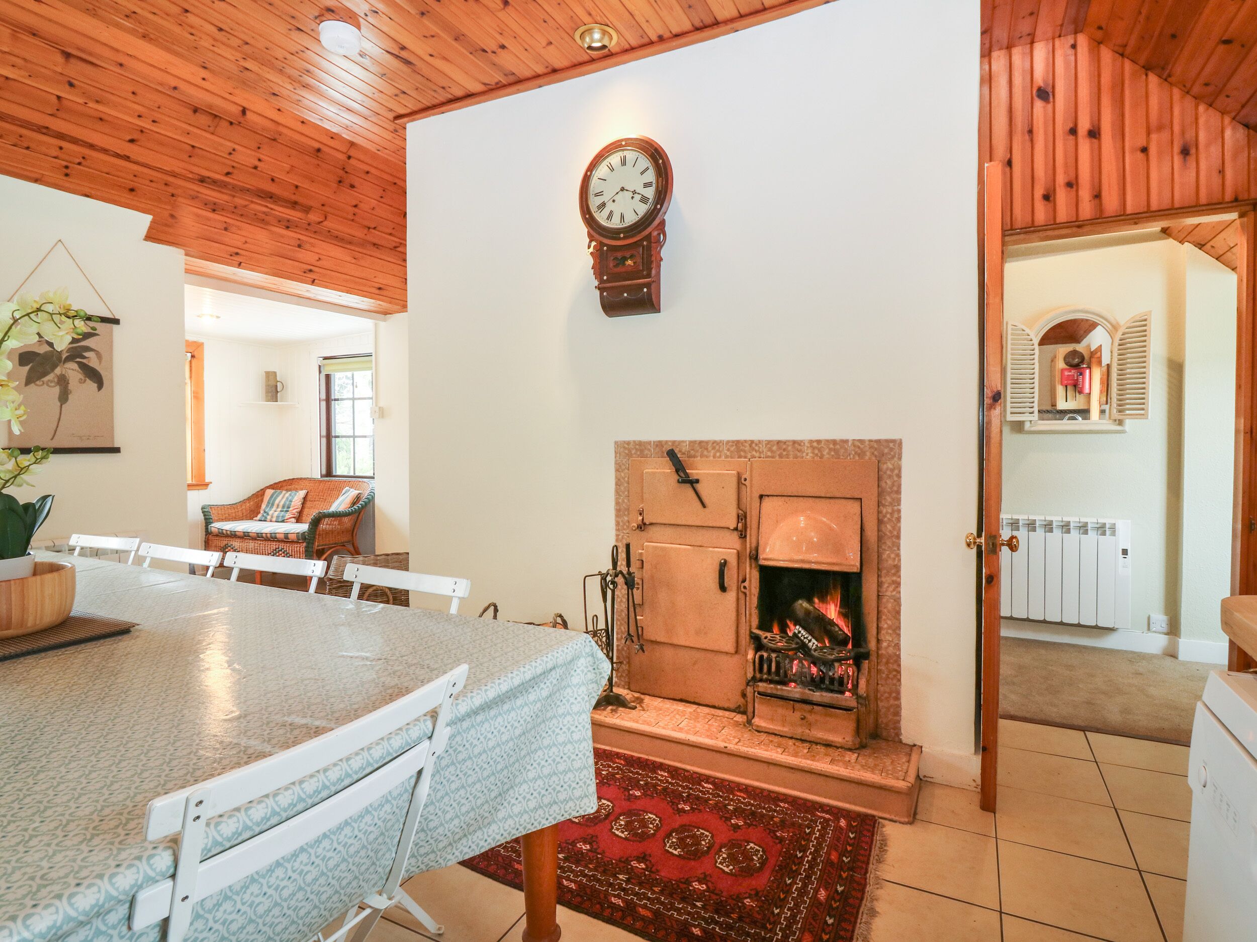 Uma sala de jantar com mesa e cadeiras de madeira, um relógio fixado na parede e uma lareira com fogo.