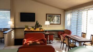 Quarto de hotel com sofá, mesa de jantar com cadeiras, televisão presa na parede e janela com persianas.