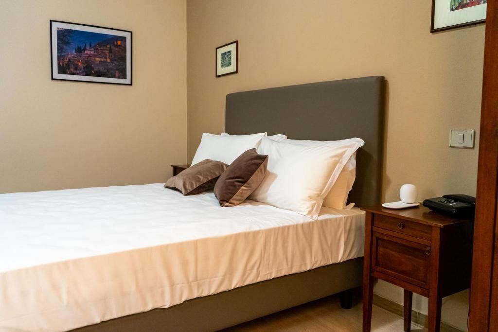 Uma cama bem arrumada com almofadas brancas e castanhas, uma mesa de cabeceira de madeira com um telefone, e duas molduras na parede.