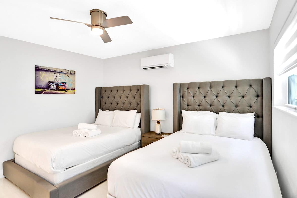 Quarto de hotel com duas camas, um ventilador de teto, um aparelho de ar condicionado e um quadro na parede.