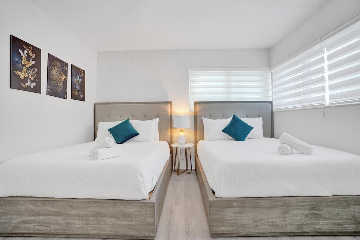 Quarto de hotel com duas camas, roupa de cama branca, almofadas verde-água, mesa de cabeceira pequena com abajur e uma obra de arte na parede.