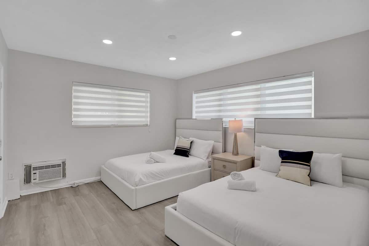 Quarto de hotel com duas camas, uma mesa de cabeceira com luminária e uma janela com persianas.