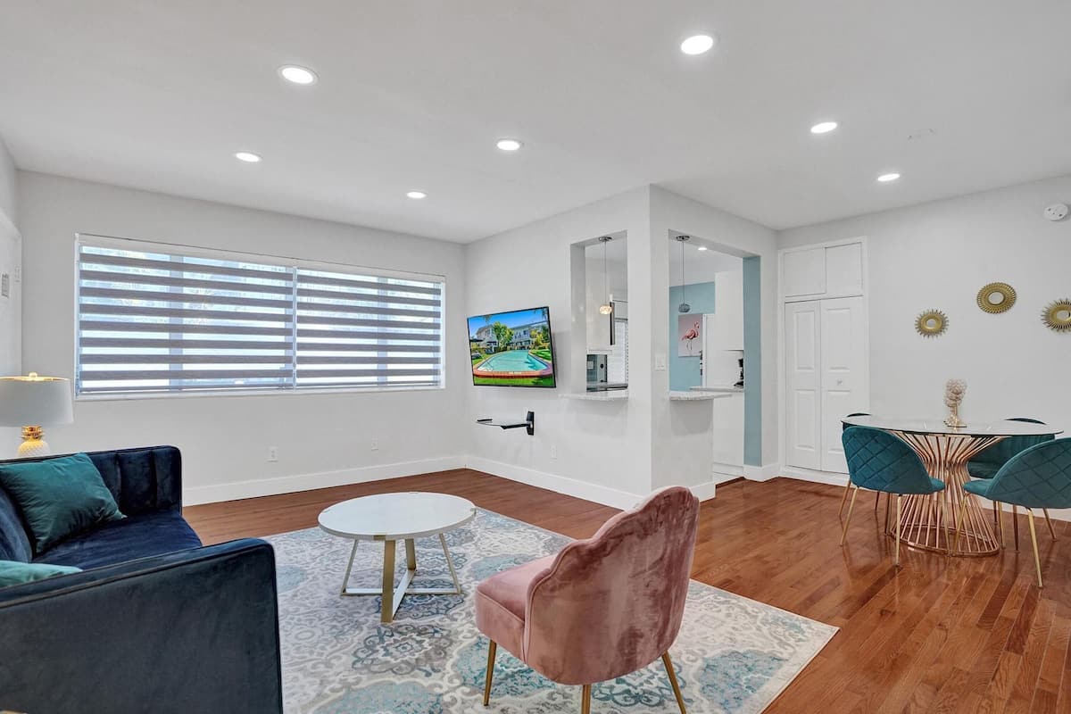Uma sala de estar moderna com um sofá azul, uma poltrona rosa, uma mesa de centro redonda e uma área de jantar com cadeiras verde-água e uma mesa dourada.