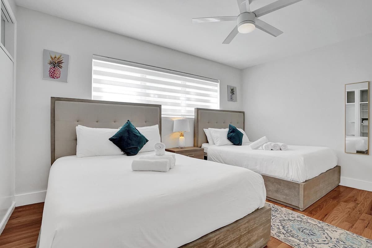Quarto de hotel com duas camas, ventilador de teto, janela com persianas e piso de madeira.