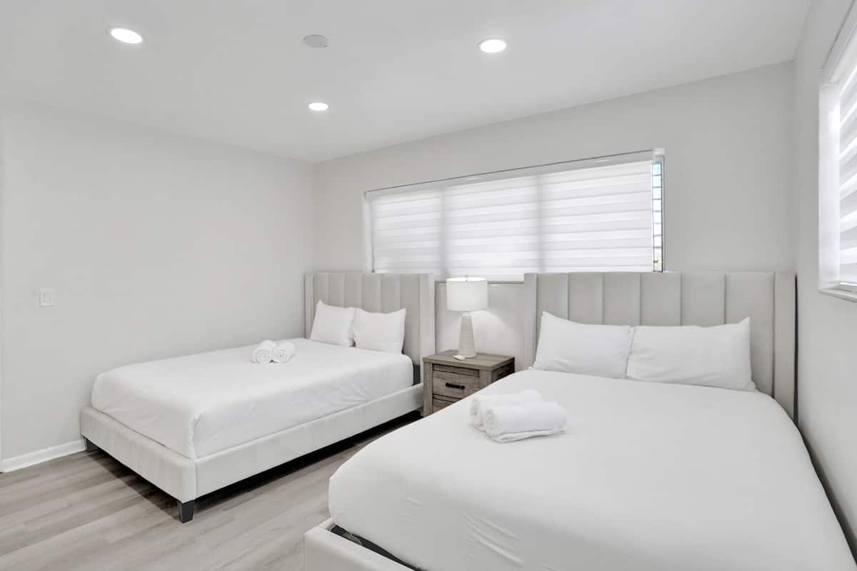 Quarto de hotel com duas camas de solteiro, mesa de cabeceira com abajur e uma janela grande com persianas.