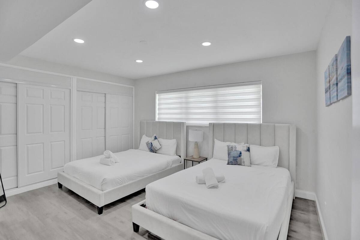 Quarto de hotel com duas camas, uma janela grande com persianas, teto branco e porta com design em painel.