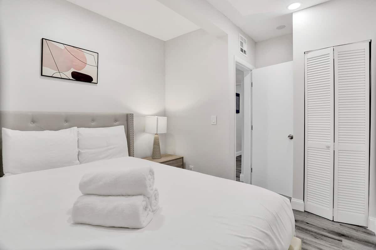 Cama bem arrumada com lençóis e toalhas brancas, mesa de cabeceira com abajur, obra de arte abstrata na parede e guarda-roupa branco.