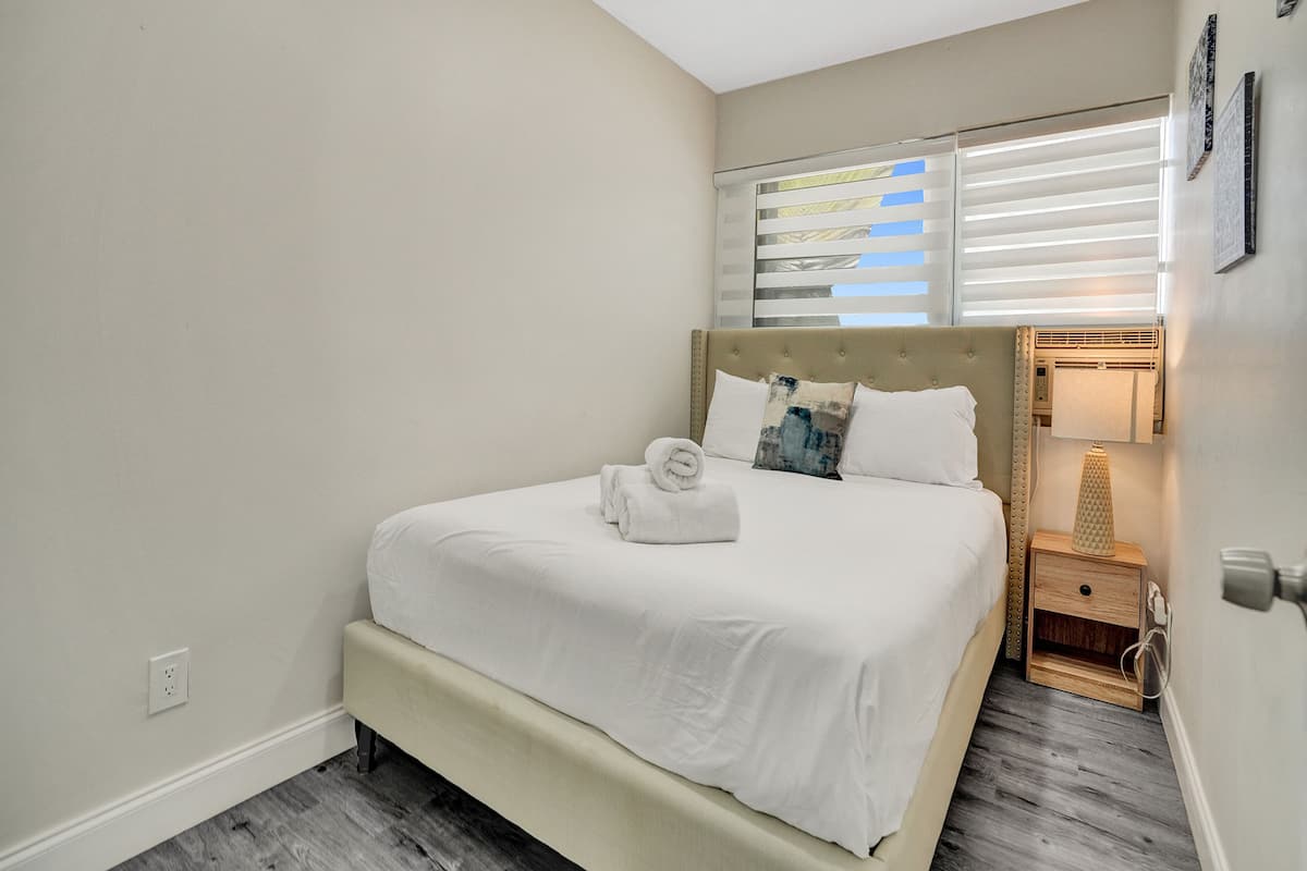 Cama bem arrumada com lençóis brancos e duas almofadas, mesa de cabeceira com abajur, e janela com persianas.