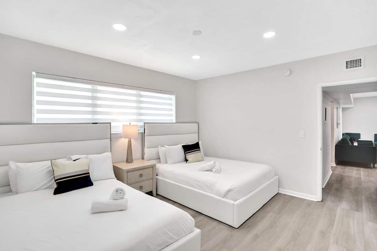 Quarto de hotel moderno com duas camas de solteiro, uma mesa de cabeceira com abajur e uma janela grande com persianas.