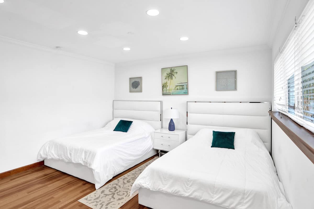 Quarto de hotel moderno com duas camas de solteiro, uma pequena mesa de cabeceira com abajur e uma janela com persianas.