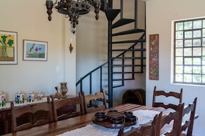 Uma sala de jantar com mesa de madeira, cadeiras, um candelabro, escada em espiral e obras de arte nas paredes.