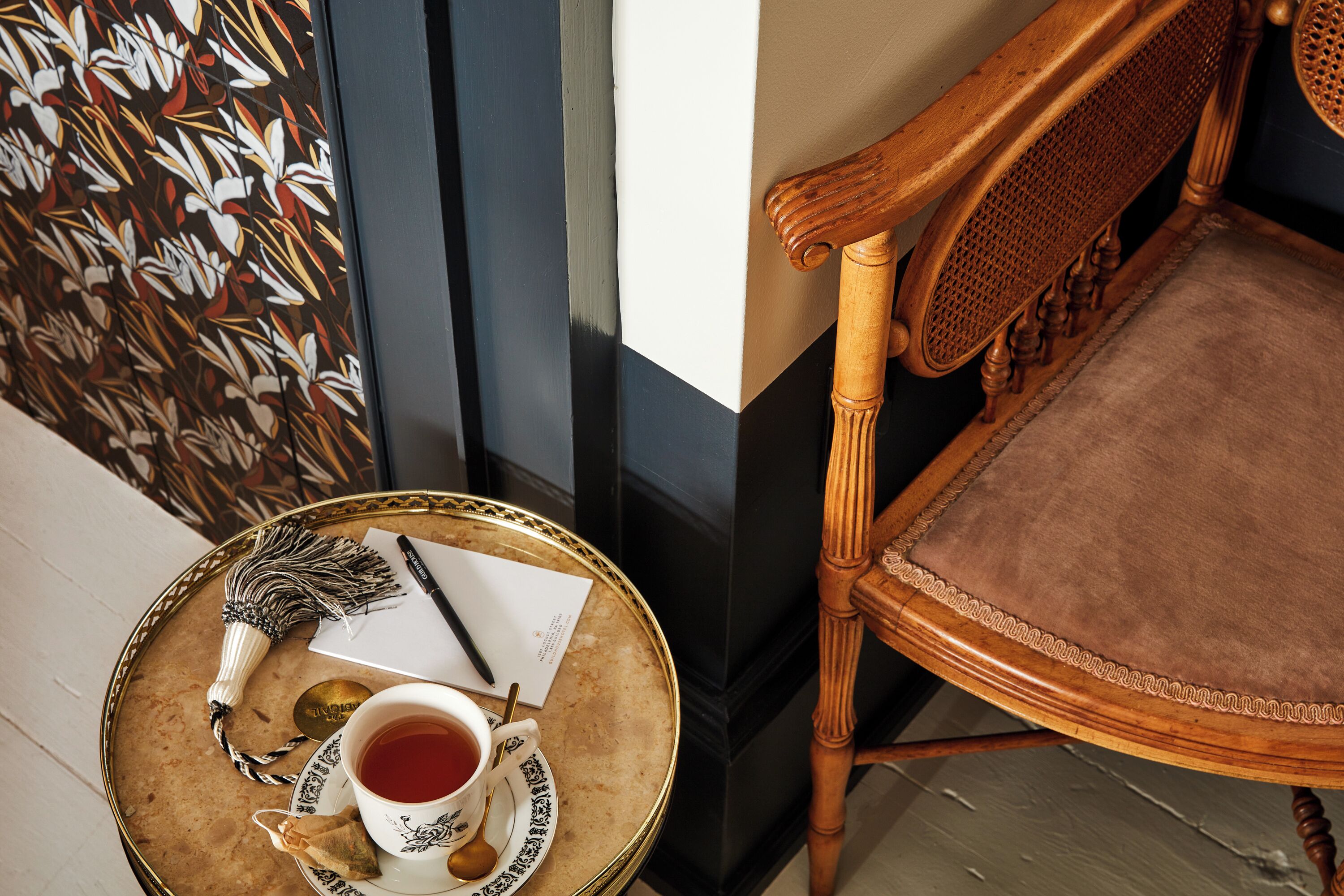 Uma cadeira de madeira com assento acolchoado, uma mesa redonda com um tabuleiro que contém uma chávena de chá, uma caneta, um caderno e um objeto decorativo.