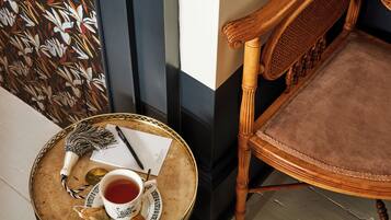 Uma cadeira de madeira com assento acolchoado, uma mesa redonda com um tabuleiro que contém uma chávena de chá, uma caneta, um caderno e um objeto decorativo.