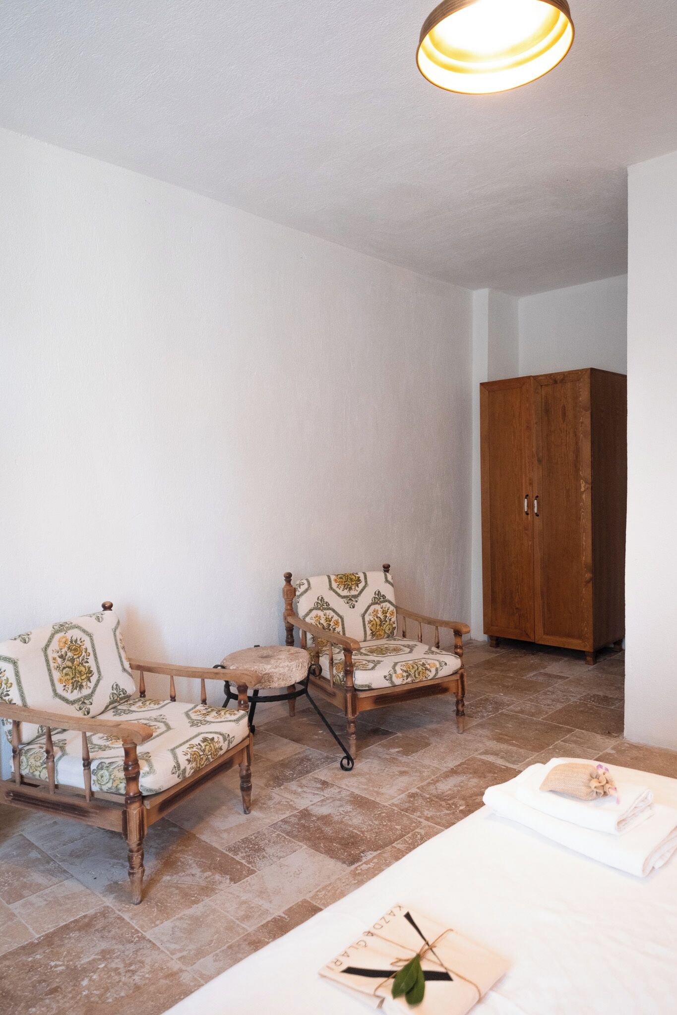 Quarto com duas cadeiras de madeira com estofamento floral, um guarda-roupa de madeira e uma cama com colcha branca e uma toalha.