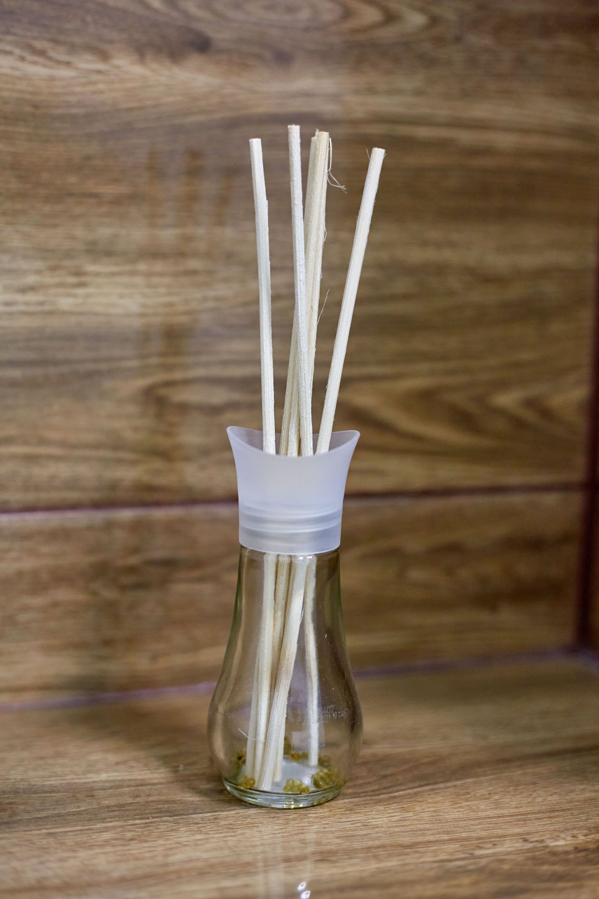 Um vaso de vidro transparente com tampa branca, contendo vários paus de junco e pequenas flores amarelas, apoiado sobre uma superfície de madeira.