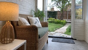 Um quarto aconchegante com um sofá de vime, uma mesa redonda com abajur, e vista para um jardim através de uma porta de vidro.