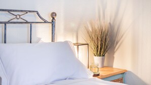 Cama bem arrumada com lençóis brancos, mesa de cabeceira com um copo de água, abajur e uma planta em vaso.