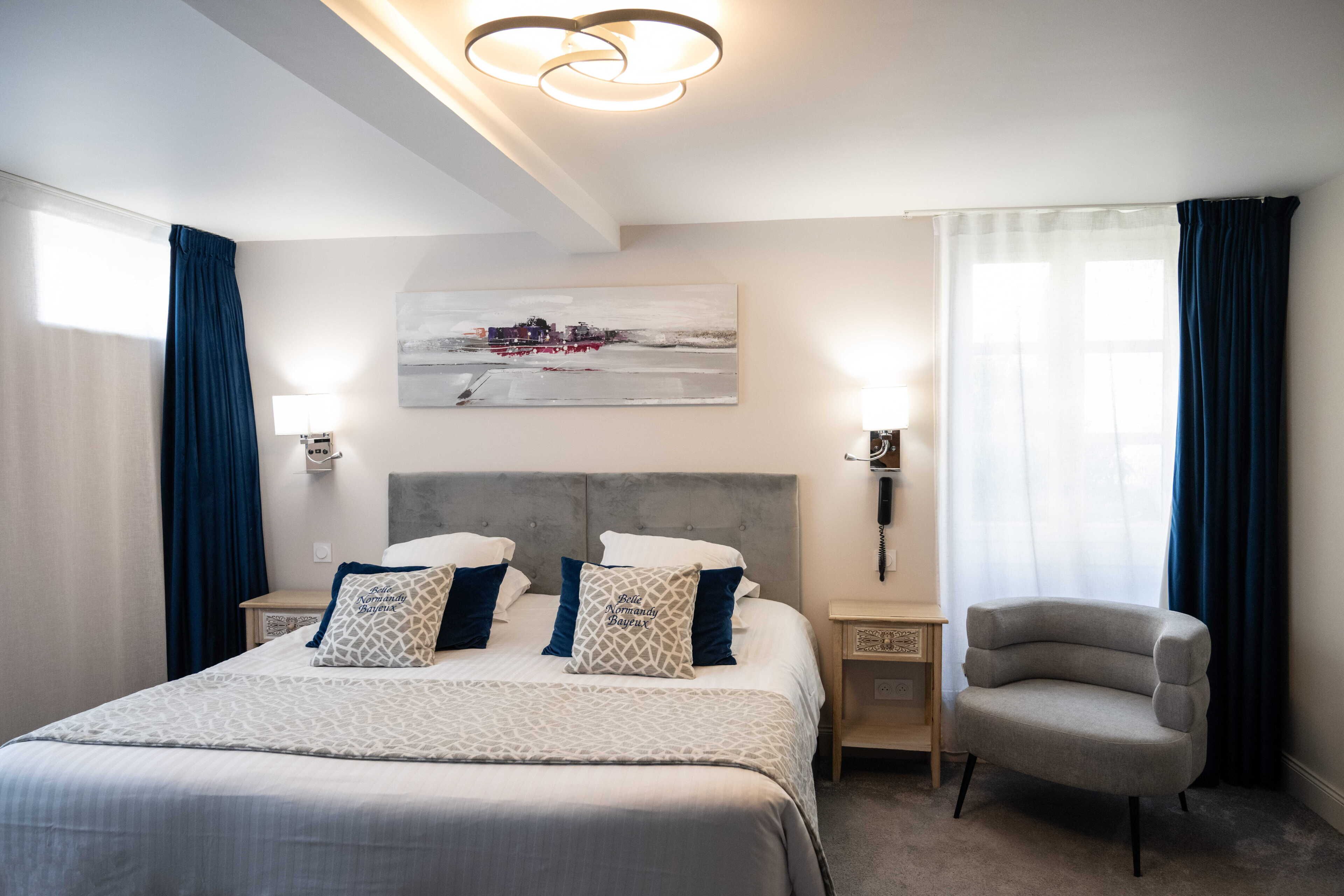 Quarto de hotel com uma cama grande, uma cadeira, mesa de cabeceira, luminária na parede e um quadro.