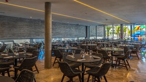 Um restaurante espaçoso com mesas redondas e cadeiras de vime, grandes janelas e vista para um parquinho colorido.