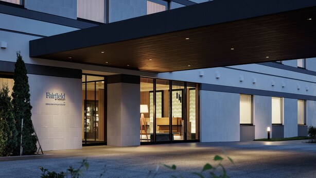 Entrada moderna de um hotel com galeria coberta, portas de vidro e uma placa com a inscrição 'Fairfield by Marriott'.