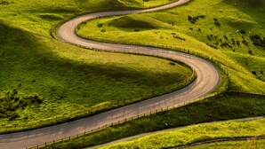 Uma estrada sinuosa por um vale verde com o nascer ou pôr do sol ao fundo.