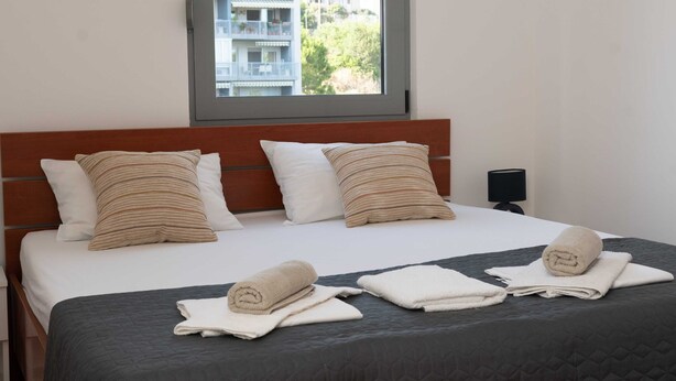 Cama bem arrumada com colcha escura, travesseiros brancos e bege, e toalhas dobradas numa janela com vista para prédios.