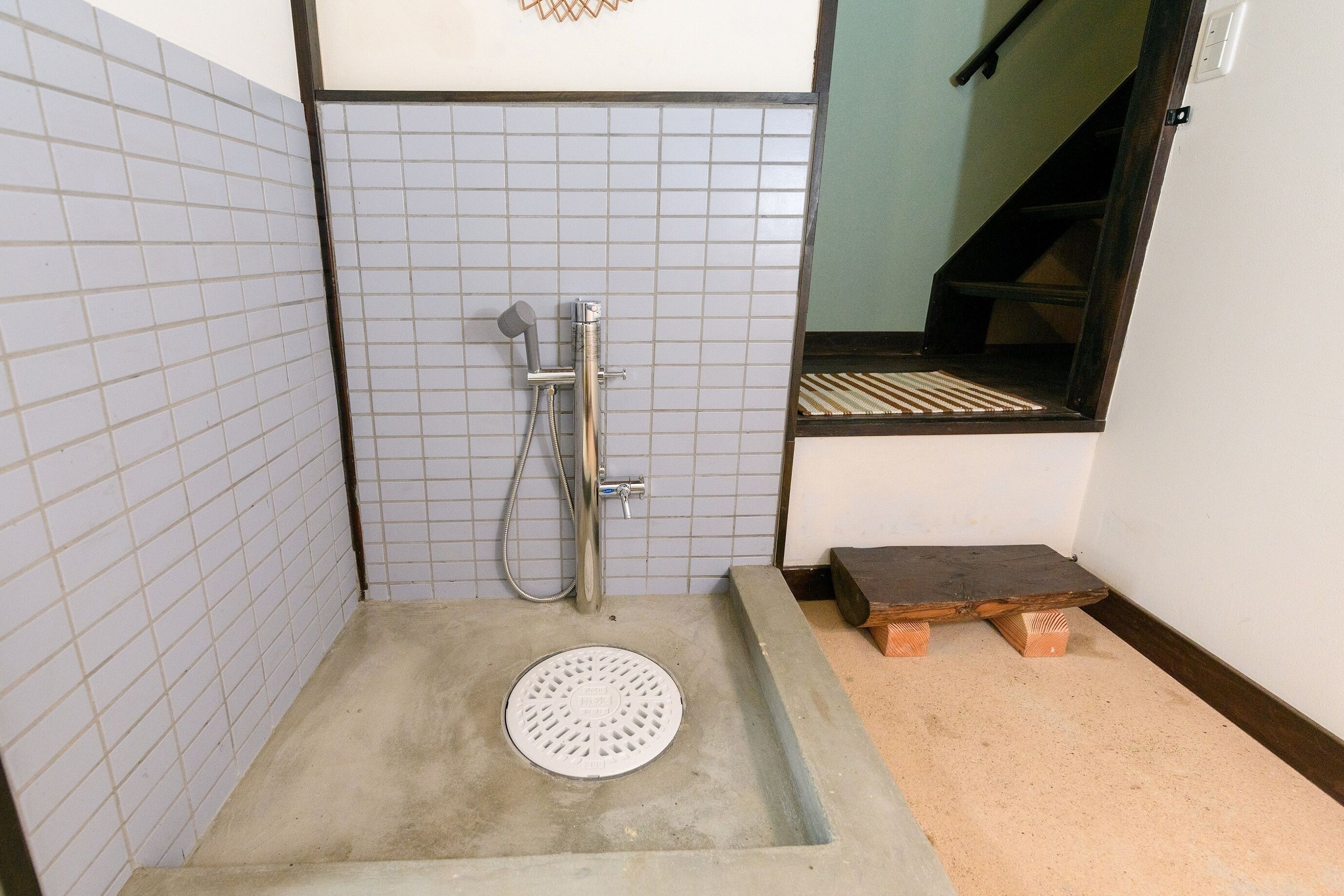 Um banheiro pequeno com azulejos, chuveiro, ralo e um banco de madeira.