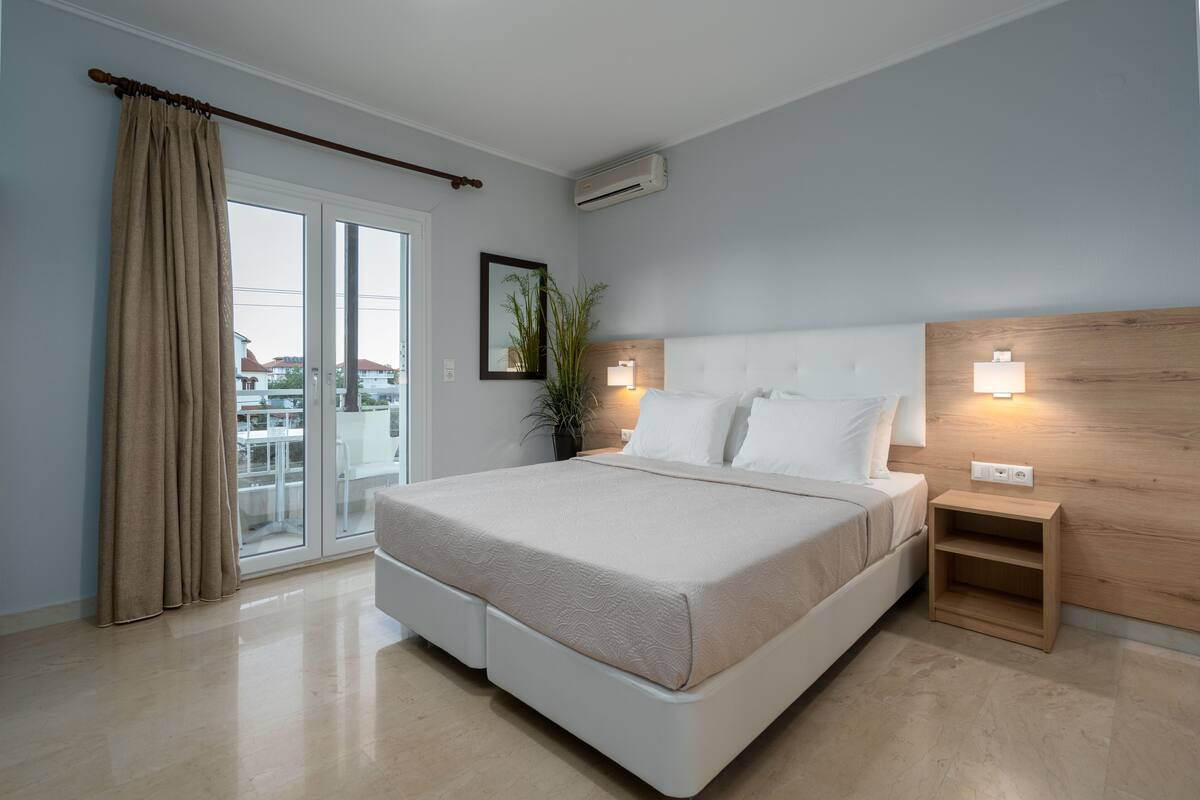 Quarto moderno com uma cama grande, portas de vidro deslizantes para uma varanda, uma pequena mesa de cabeceira e ar-condicionado fixado na parede.