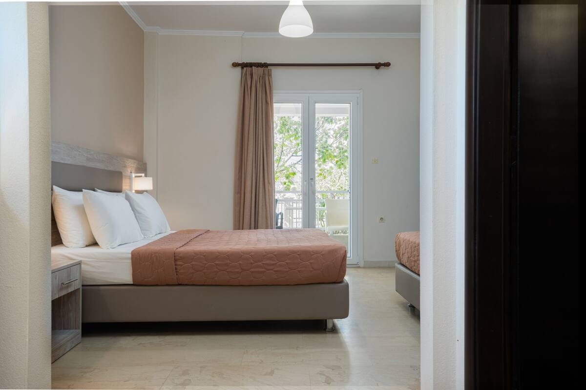 Quarto com cama, mesas de cabeceira, janela com cortinas e vista para o verde lá fora.