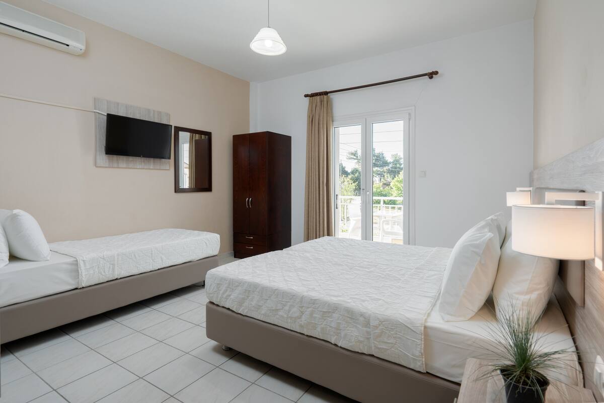 Quarto de hotel com duas camas, televisão, guarda-roupa e varanda com cortinas.