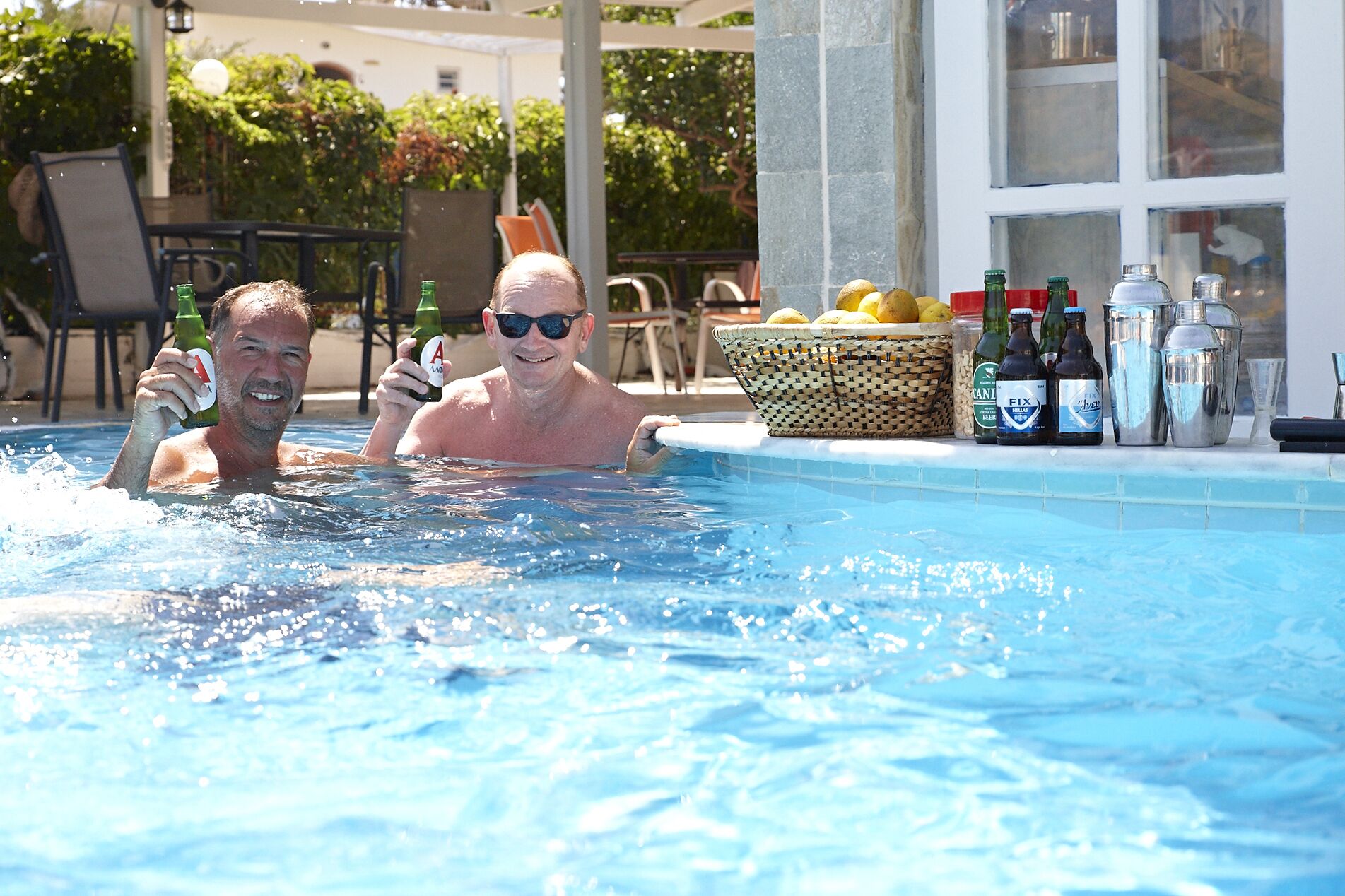 Uma piscina com duas pessoas, uma cesta de vime com frutas e garrafas de cerveja na beira da piscina.